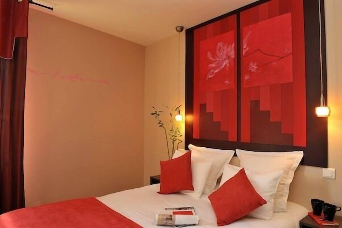 Quarto de hotel com cama, almofadas vermelhas e brancas, mesa de cabeceira com abajur e uma parede com arte abstrata vermelha.