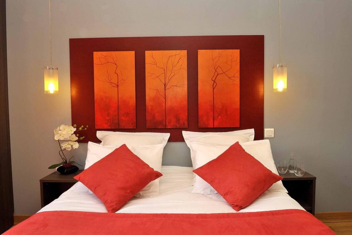 Quarto de hotel com uma cama grande, duas almofadas vermelhas, um vaso com flores brancas e três quadros na parede.