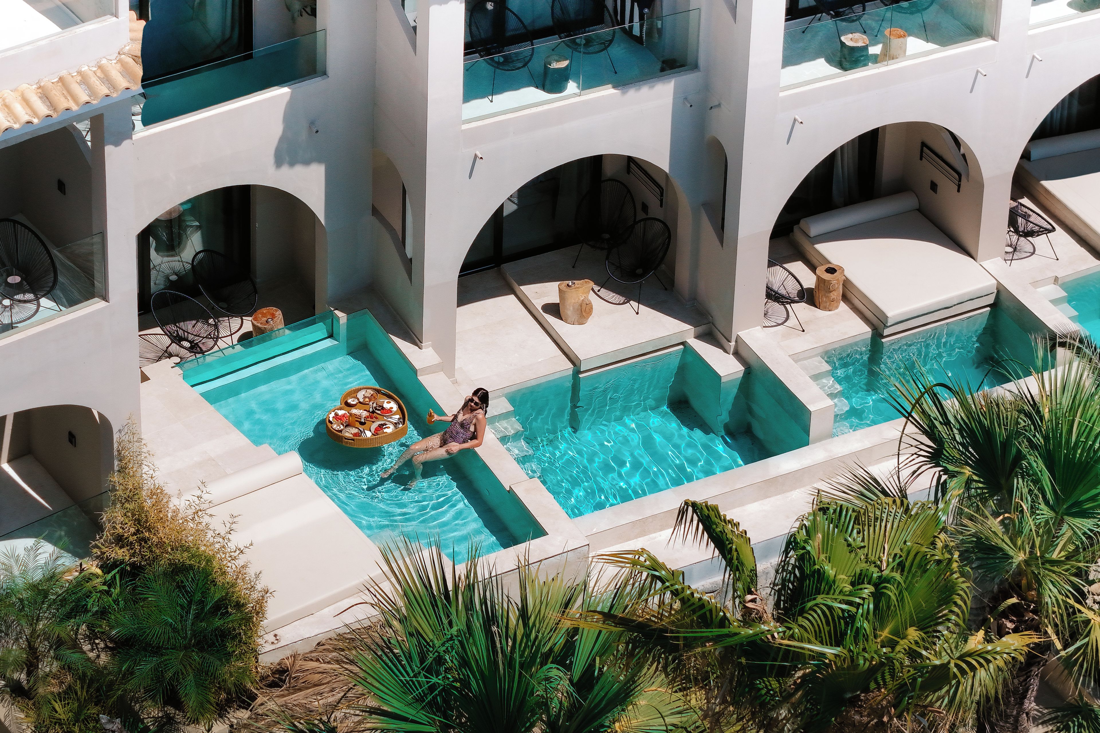 Uma área de piscina com várias seções, incluindo uma piscina central e piscinas menores, cercada por espreguiçadeiras e um espaço para refeições.