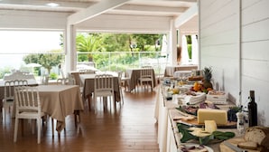 Restaurante com mesas e cadeiras, um buffet com diversos pratos e vista para o verde lá fora.