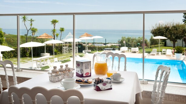 Mesa de pequeno-almoço com vista para a piscina e o mar, com mobiliário branco e guarda-sóis.