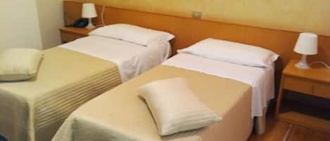 Quarto de hotel com duas camas de solteiro, cada uma com almofada e mesa de cabeceira com abajur.