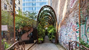 Uma passagem urbana com uma escultura de metal, um muro de tijolos coberto por grafites e um banco de madeira.