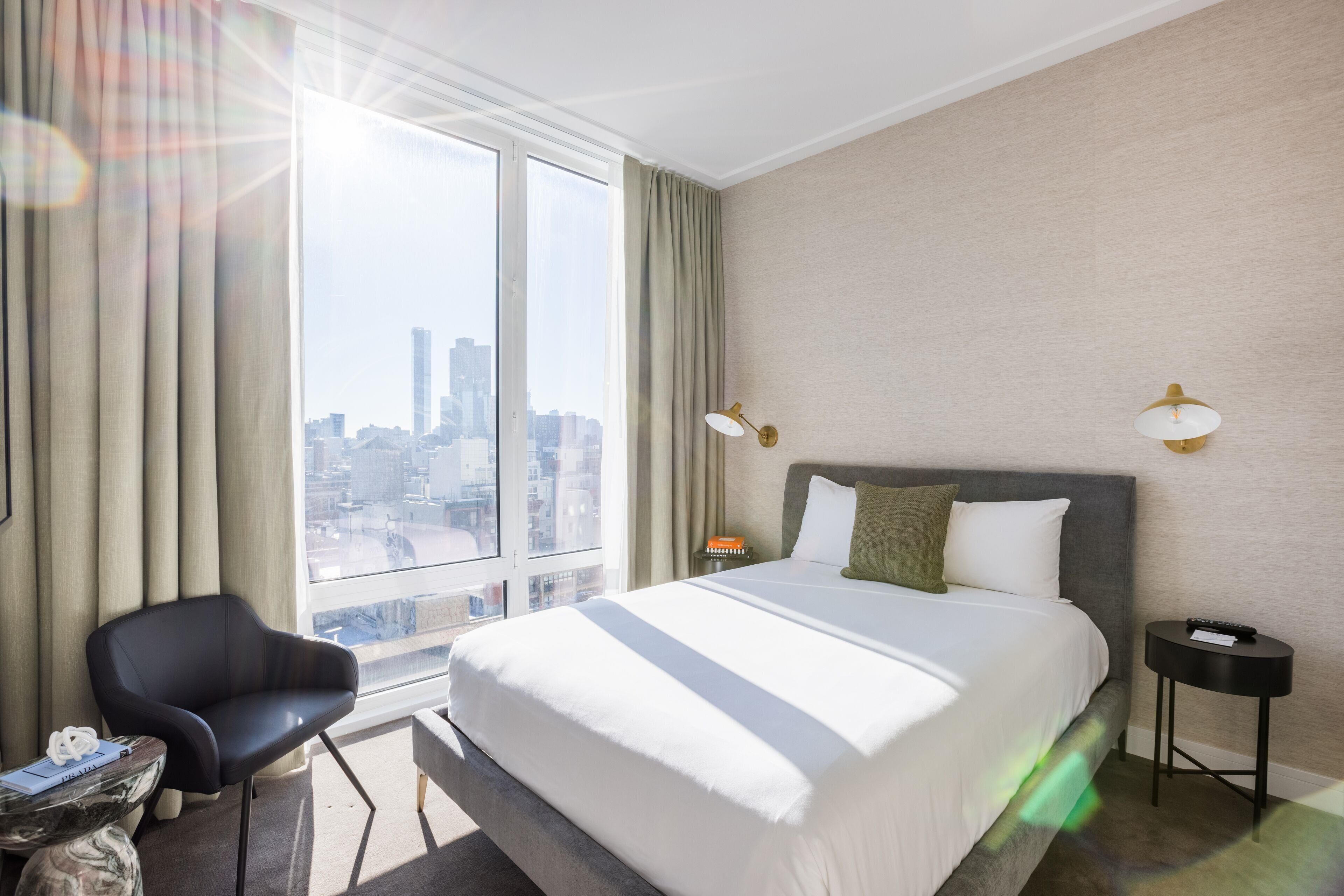 Quarto de hotel com uma cama grande, uma cadeira, uma mesinha de cabeceira e vista para a cidade.