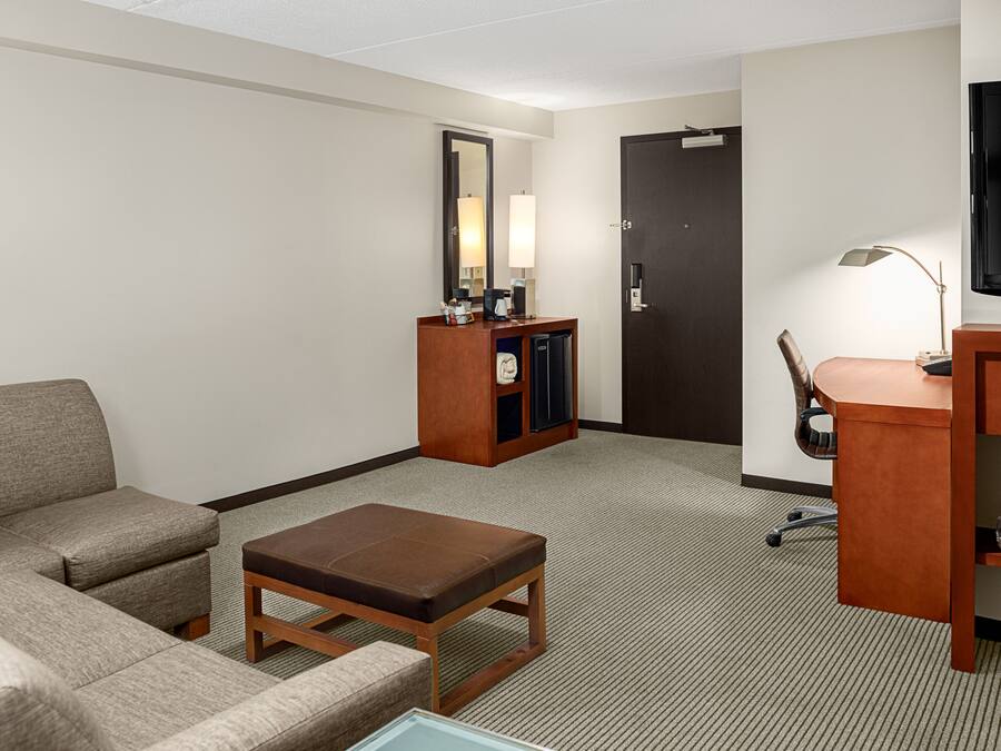 Quarto de hotel com sofá, mesa pequena, escrivaninha com cadeira, televisão, espelho e porta.