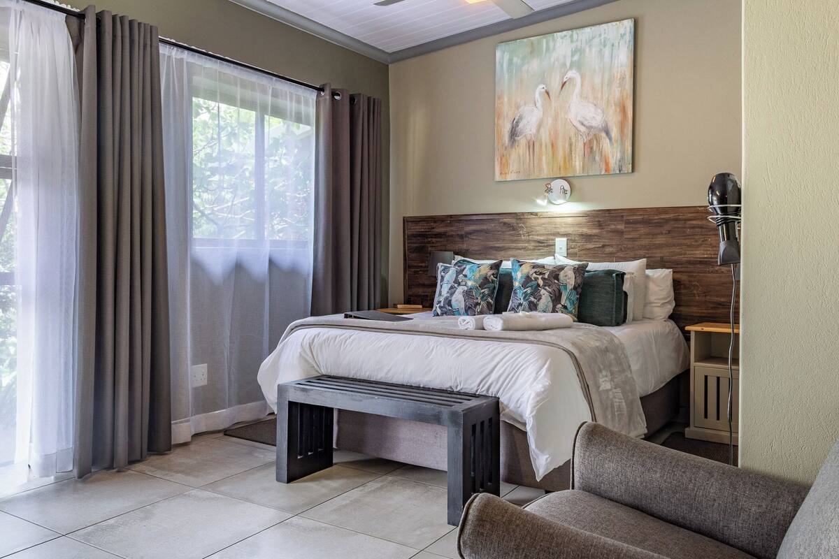 Quarto com uma cama grande, um banquinho, uma mesa de cabeceira e um quadro na parede.