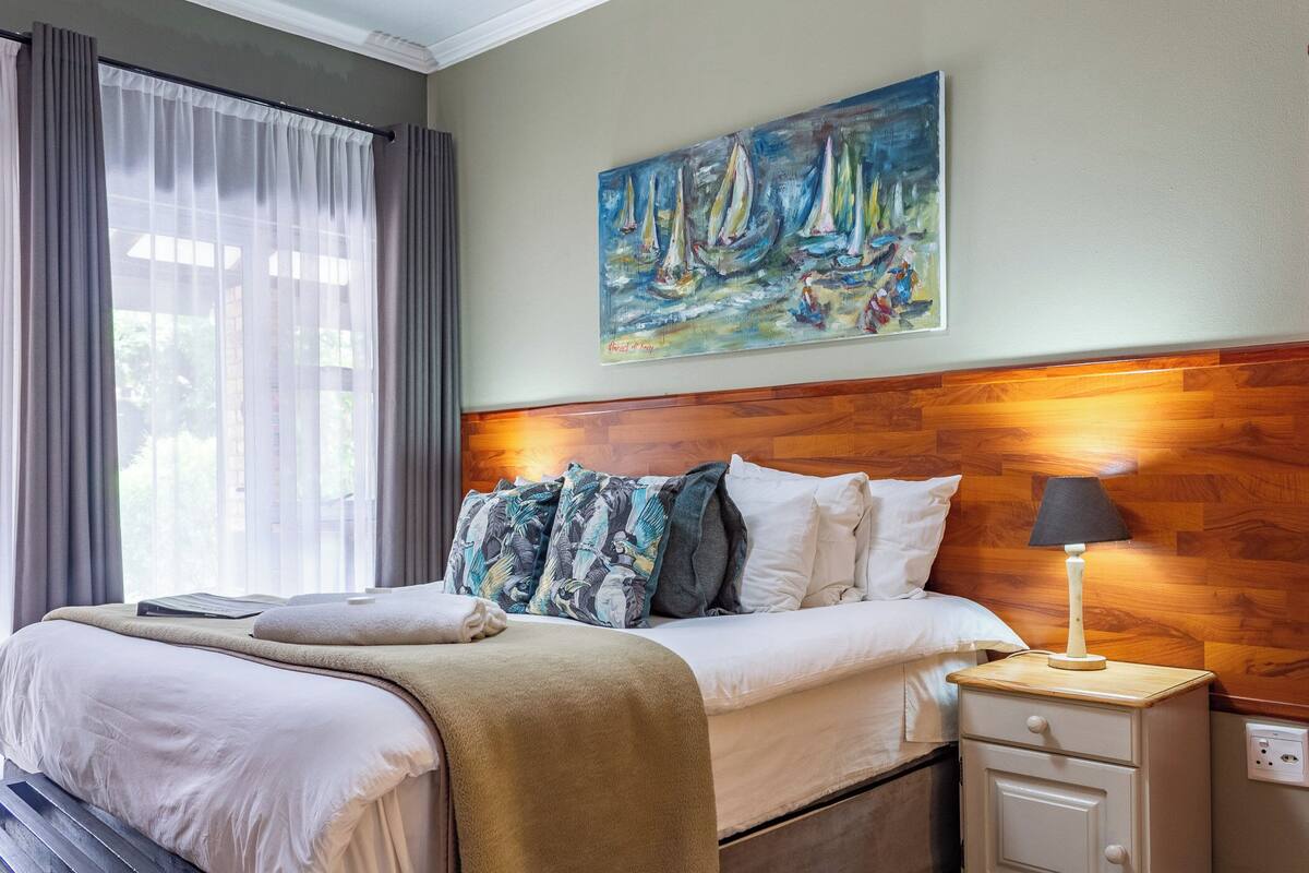 Uma cama bem arrumada com um cobertor bege, uma mesa de cabeceira com abajur, e um quadro com barcos à vela na parede.