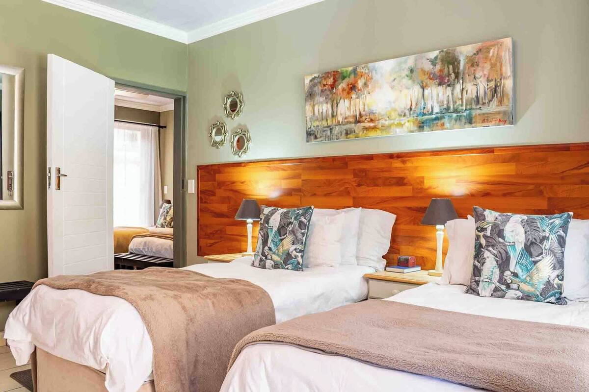 Quarto de hotel com duas camas, cabeceira de madeira, um quadro e uma porta que dá para outro cômodo.