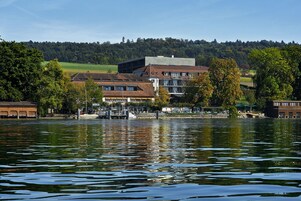 Vista para um lago com edifícios, um cais e uma colina arborizada ao fundo.
