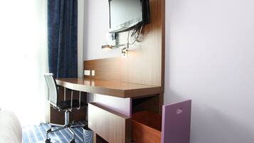 Quarto de hotel compacto com escrivaninha de madeira, uma cadeira, televisão fixada na parede e um móvel de armazenamento roxo.