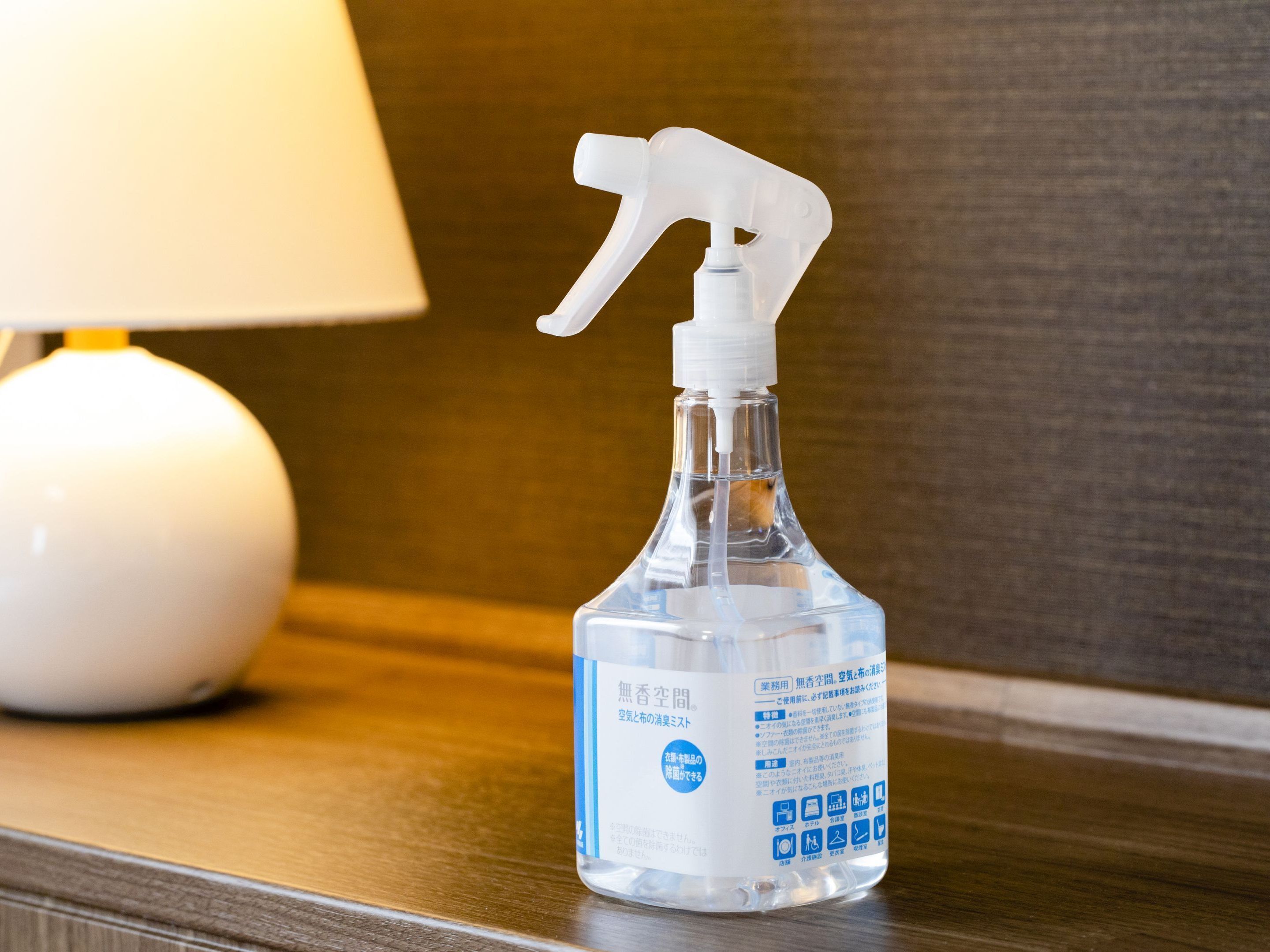 Frasco de spray de plástico transparente com bocal branco, apoiado em superfície de madeira ao lado de um abajur branco.