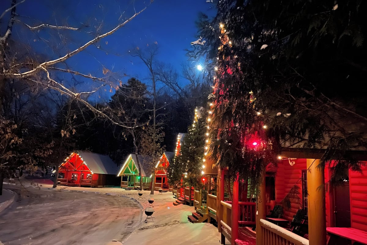 Rua coberta de neve, com edifícios iluminados com luzes festivas coloridas e um céu noturno límpido.