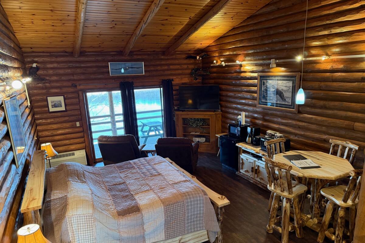 Quarto de cabana de madeira com cama, mesa de jantar, cadeiras, televisão e lareira.