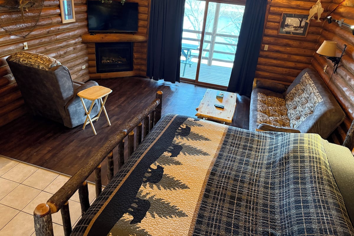 Interior acolhedor de uma cabana de toras, com lareira, televisão e uma janela ampla com vista para um deck.
