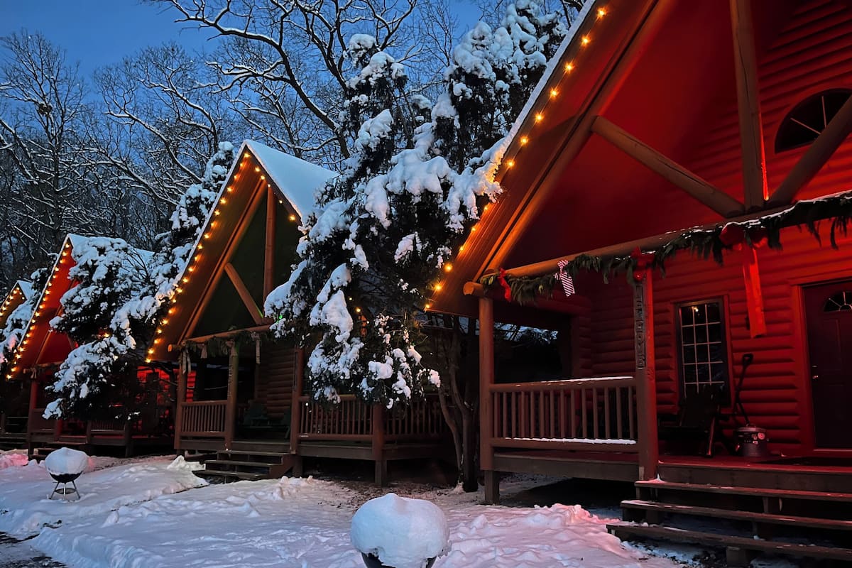Uma cabana de tronco com varanda, árvores cobertas de neve e luzes de Natal.