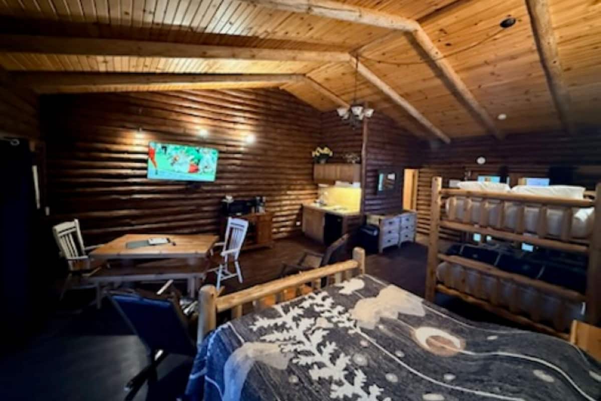 Uma cabana acolhedora com interior de madeira, uma cama com um cobertor estampado, uma escrivaninha com uma cadeira e uma televisão fixada na parede.