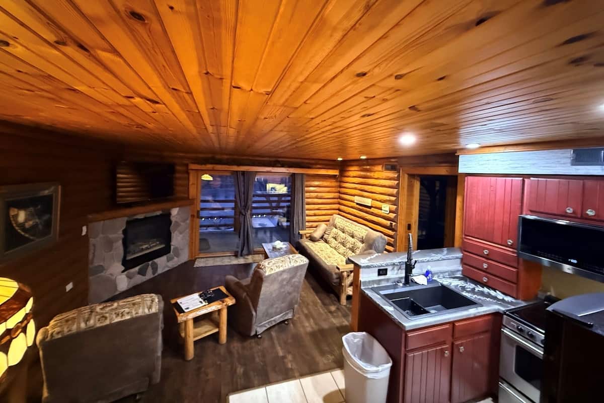 Interior acolhedor de uma cabana, com teto de madeira, lareira, sofá, mesa de jantar e área de cozinha.