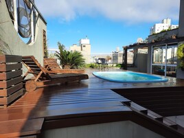 Uma área de piscina no terraço com espreguiçadeiras de madeira, uma piscina pequena e vista para edifícios ao redor.