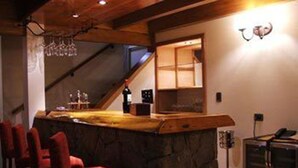 Uma área de bar com balcão de pedra, banquetas vermelhas, um candelabro e uma escada que leva a um quarto com uma janela grande.