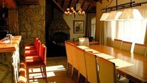 Uma sala de jantar com uma longa mesa de madeira e cadeiras vermelhas, uma parede de pedra e uma lareira.