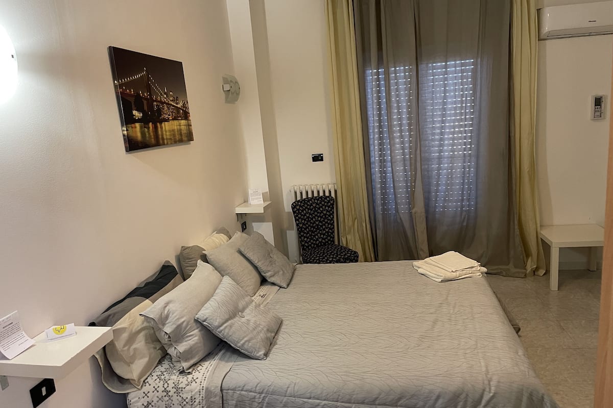 Quarto bem organizado com uma cama, travesseiros, uma cadeira, uma mesinha e ar-condicionado fixado na parede.