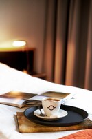 Uma chávena de café num tabuleiro com uma colher, um livro aberto e uma cama com lençóis brancos.
