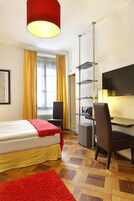 Quarto de hotel com cama, escrivaninha, cadeira, televisão, prateleira, radiador e janela com cortinas.