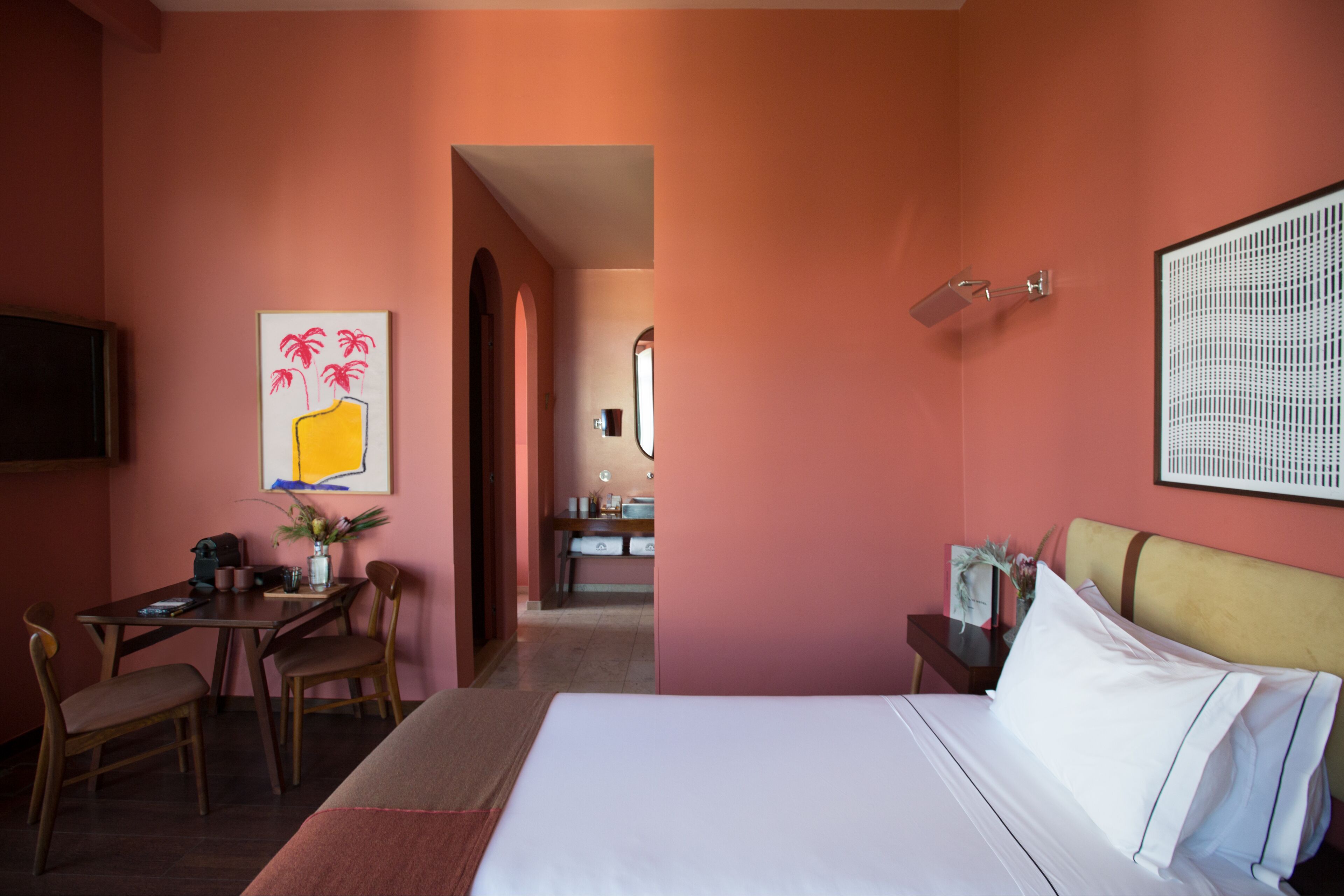 Quarto de hotel com uma cama, uma escrivaninha com cadeiras, uma televisão e um quadro na parede.