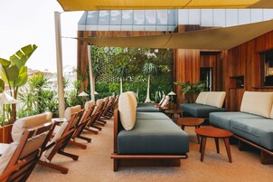Uma área de estar moderna ao ar livre, com mobiliário de madeira e sofá verde, cercada por plantas e um painel de madeira.
