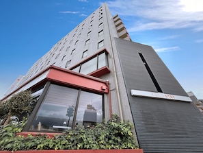 Um edifício moderno com uma placa de destaque 'KOKO STAY', janelas grandes e um detalhe vermelho na fachada.