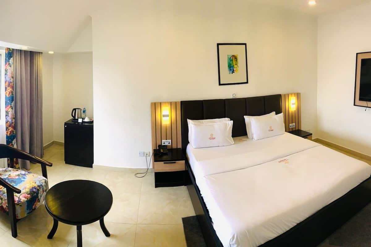 Quarto de hotel com uma cama grande, uma mesa com cadeira, uma janela com cortinas e uma mesa de cabeceira com luminária.