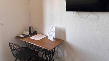 Quarto pequeno com uma escrivaninha de madeira, uma cadeira preta, uma TV de tela plana fixada na parede e uma chaleira sobre a escrivaninha.