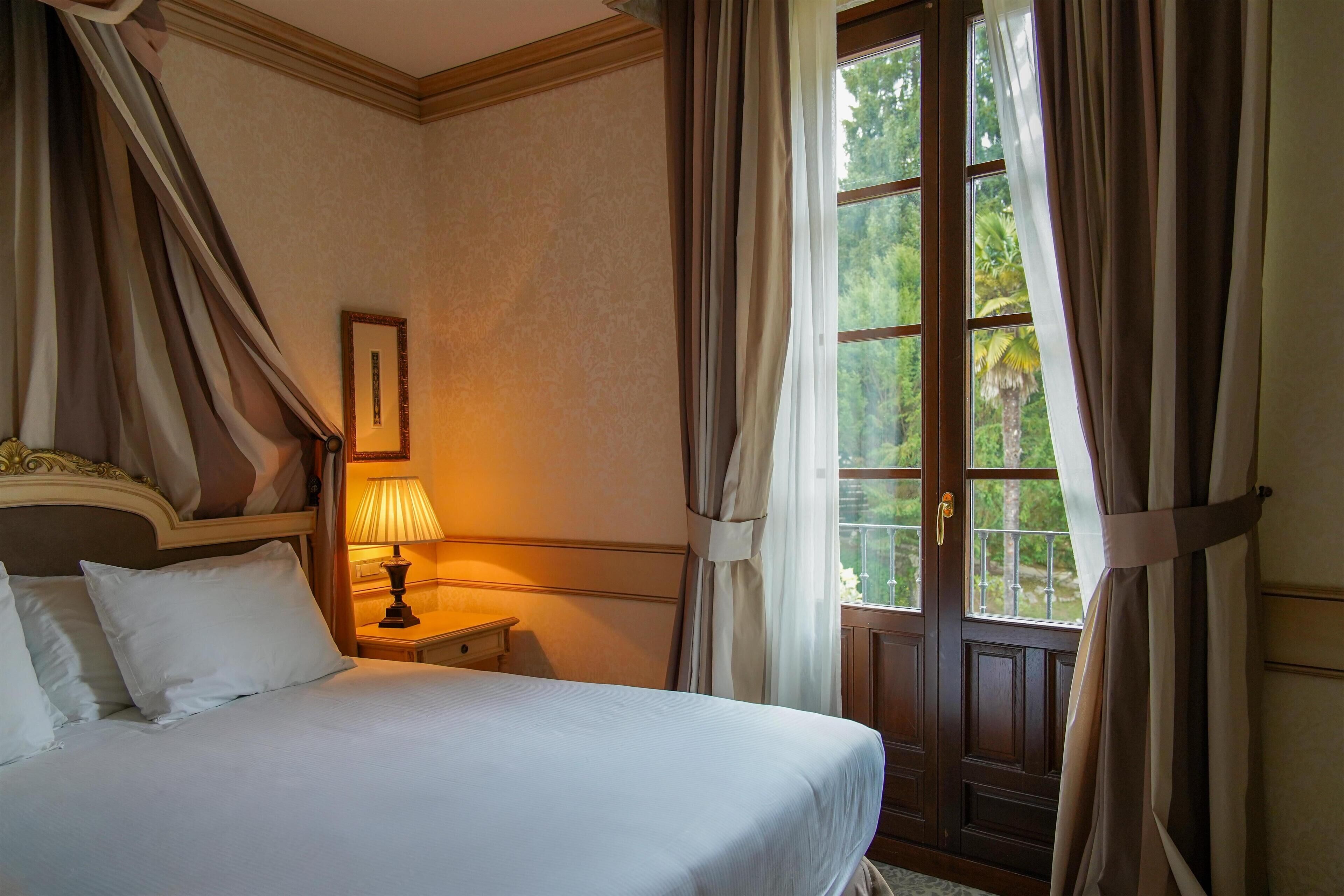 Quarto de hotel com uma cama grande, uma mesa de cabeceira com abajur e vista para o verde pela janela.