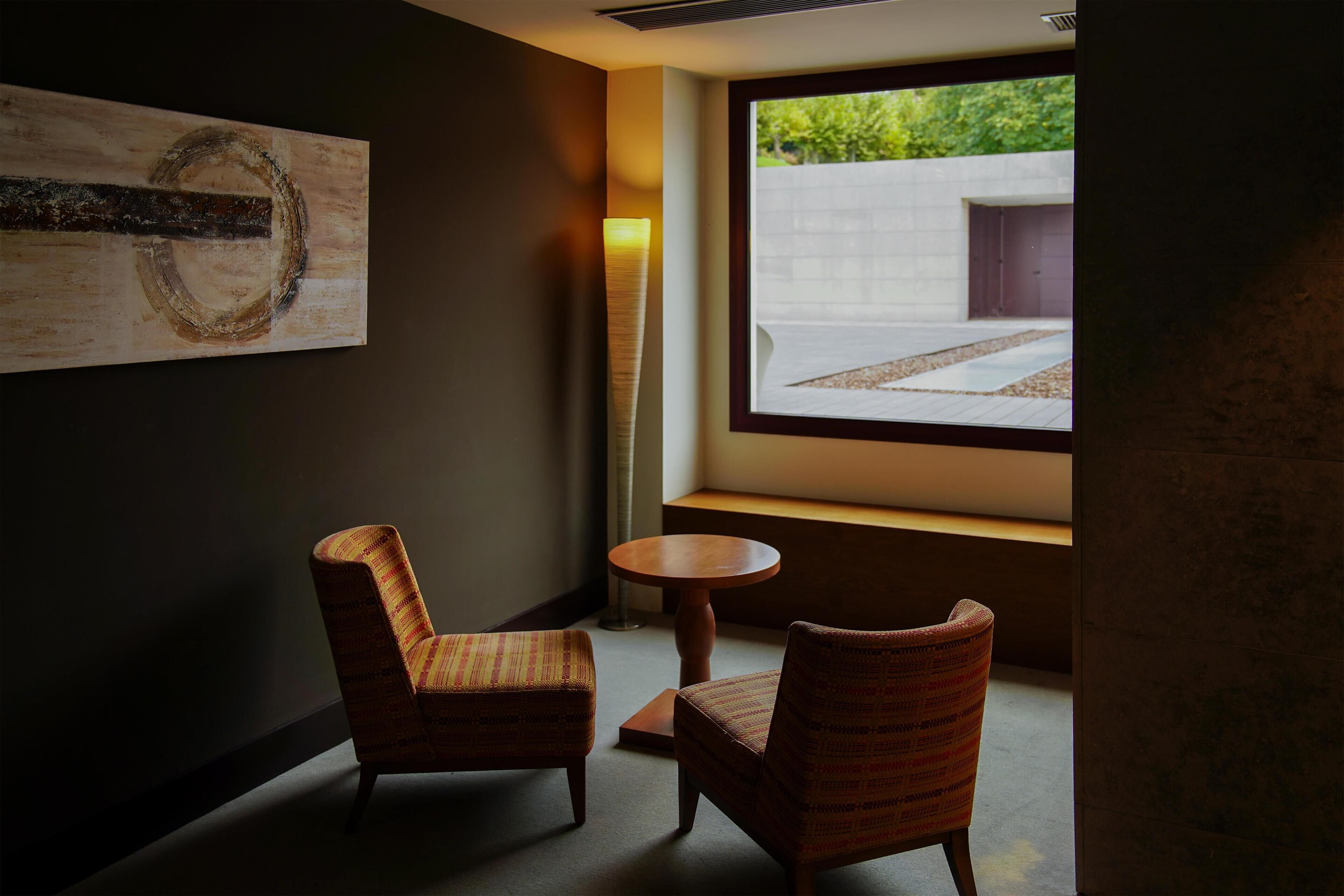 Quarto moderno com duas poltronas, uma mesinha redonda e uma janela grande que oferece vista para um edifício e vegetação.