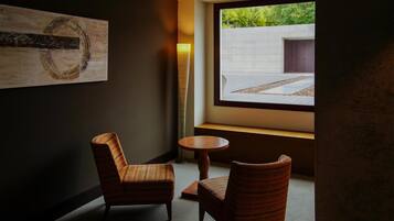 Quarto moderno com duas poltronas, uma mesinha redonda e uma janela grande que oferece vista para um edifício e vegetação.