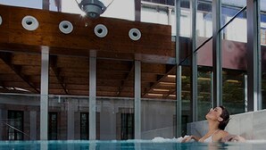 Uma pessoa relaxando em uma piscina interna transparente, com teto de madeira e janelas amplas.