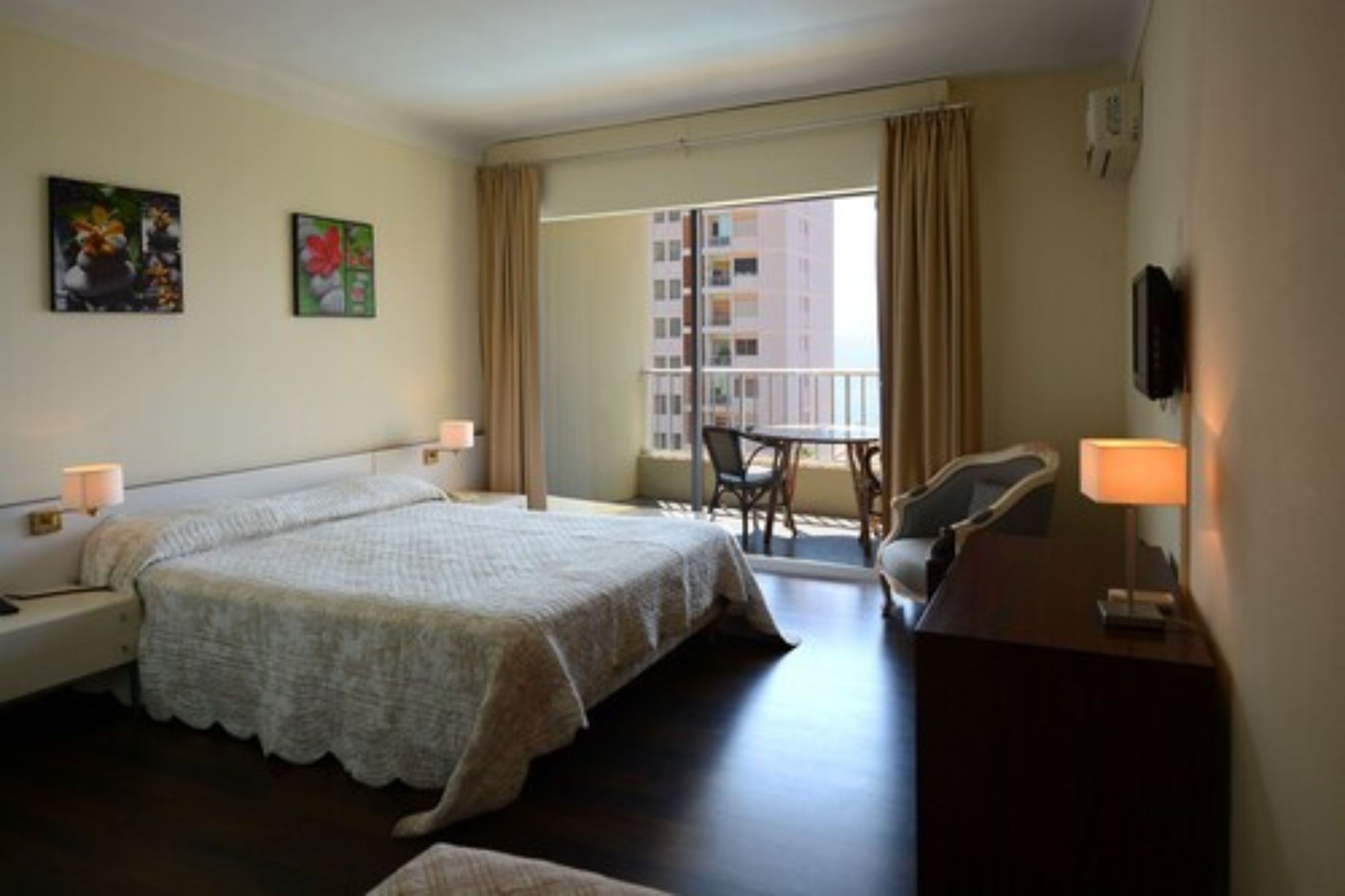 Quarto de hotel com uma cama grande, uma escrivaninha com cadeira, uma mesa pequena e uma varanda com vista para prédios.