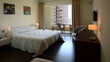 Quarto de hotel com uma cama grande, uma escrivaninha com cadeira, uma mesa pequena e uma varanda com vista para prédios.