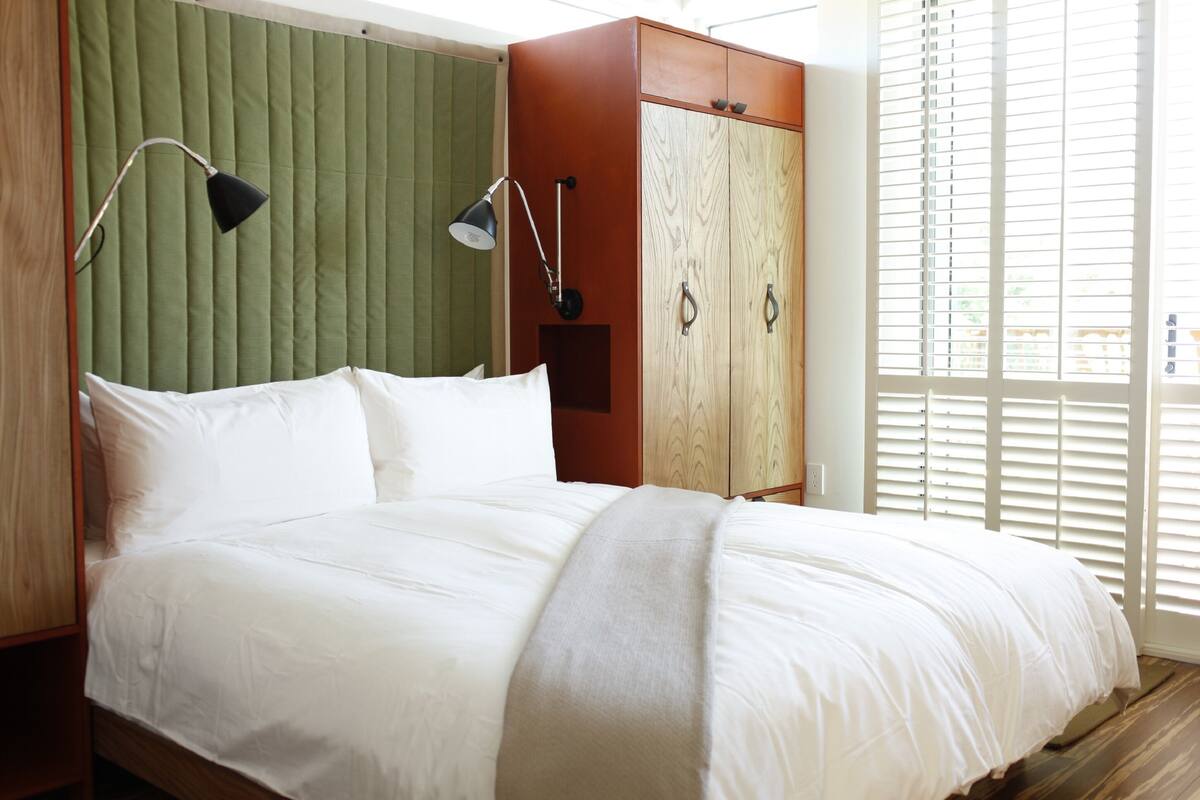 Uma cama bem arrumada com lençóis brancos, cabeceira verde e guarda-roupa de madeira com duas portas.