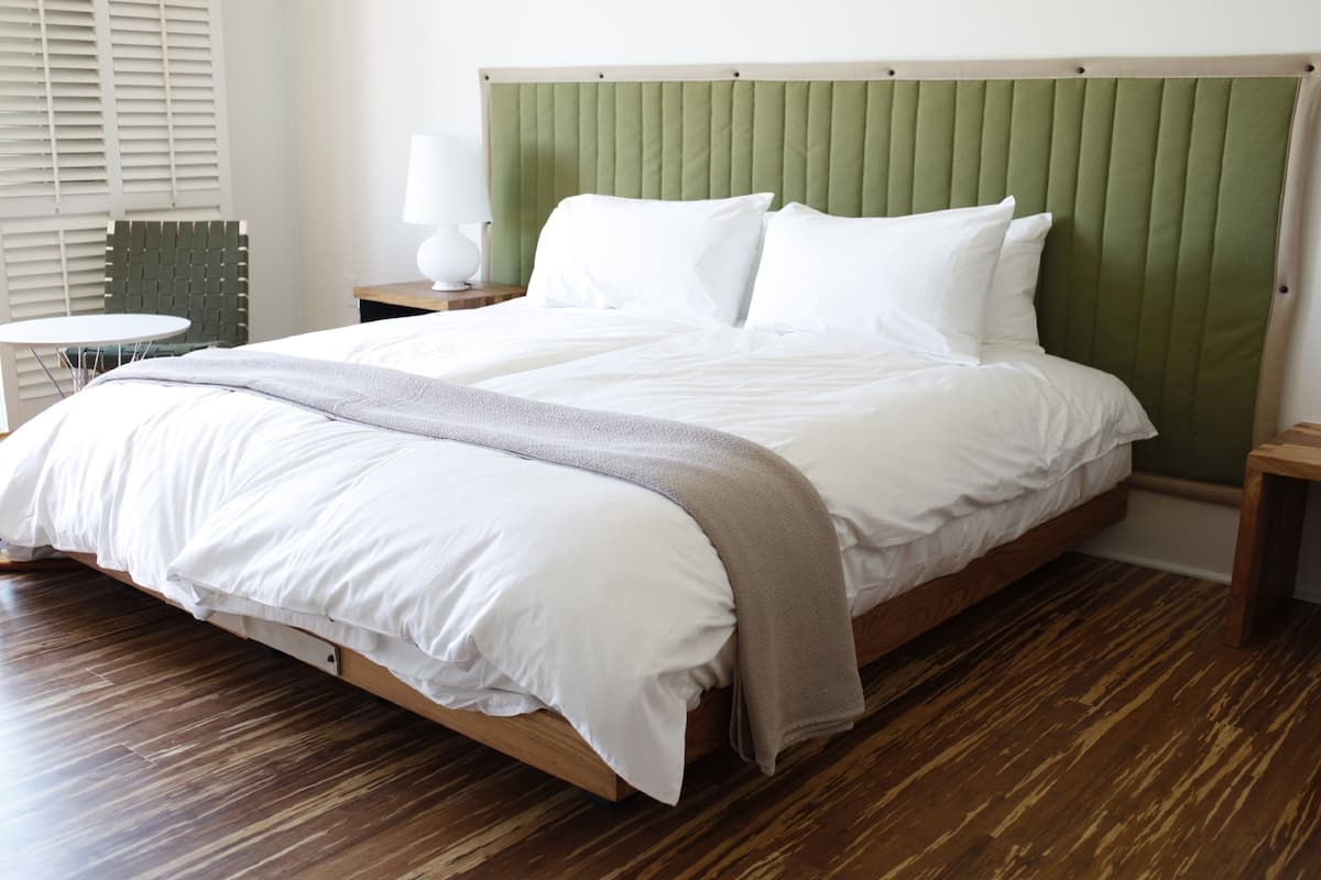 Cama bem arrumada com lençóis brancos e manta cinza, cabeceira estofada verde e chão de madeira.