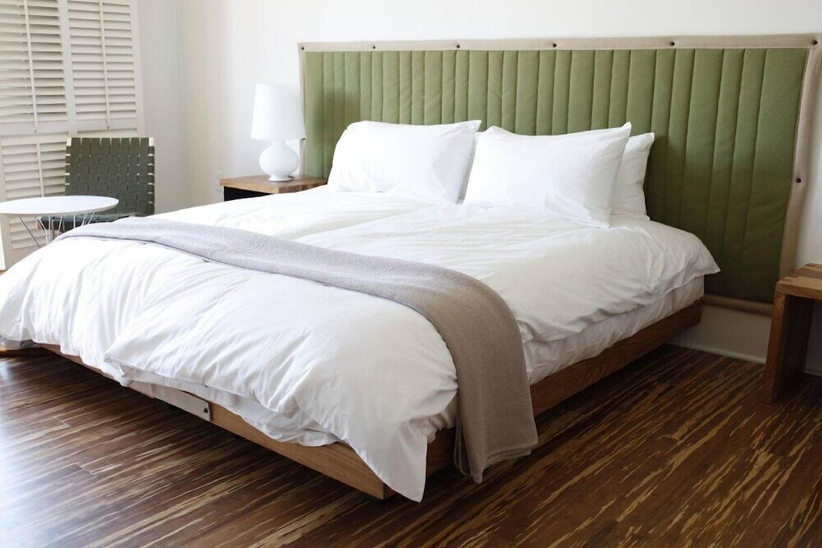Uma cama bem arrumada com estrutura de madeira, cabeceira estofada em verde e roupa de cama branca.