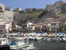 Um porto com barcos atracados, edifícios com telhados vermelhos e uma encosta rochosa ao fundo.