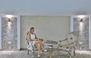 Uma mulher sentada a uma mesa redonda num quarto com paredes de mármore e duas cadeiras com estofados florais.