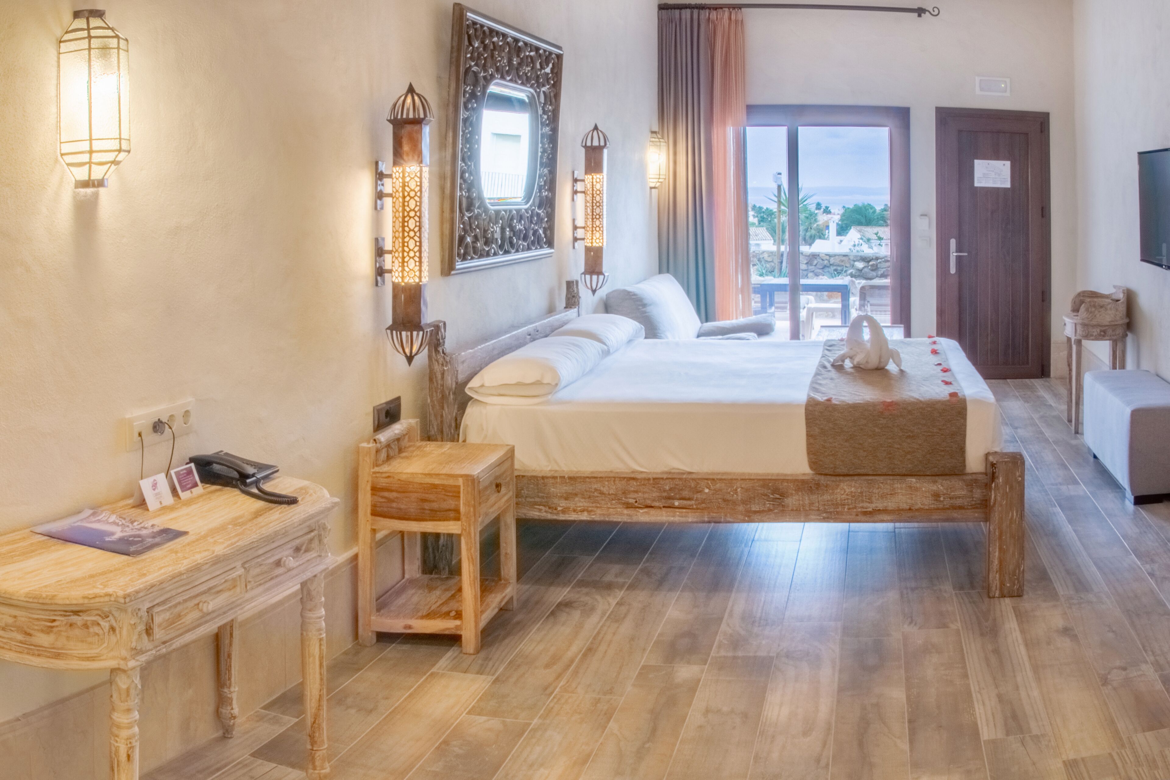 Quarto de hotel com uma cama grande, uma mesa de cabeceira de madeira, uma escrivaninha com telefone e uma televisão na parede.