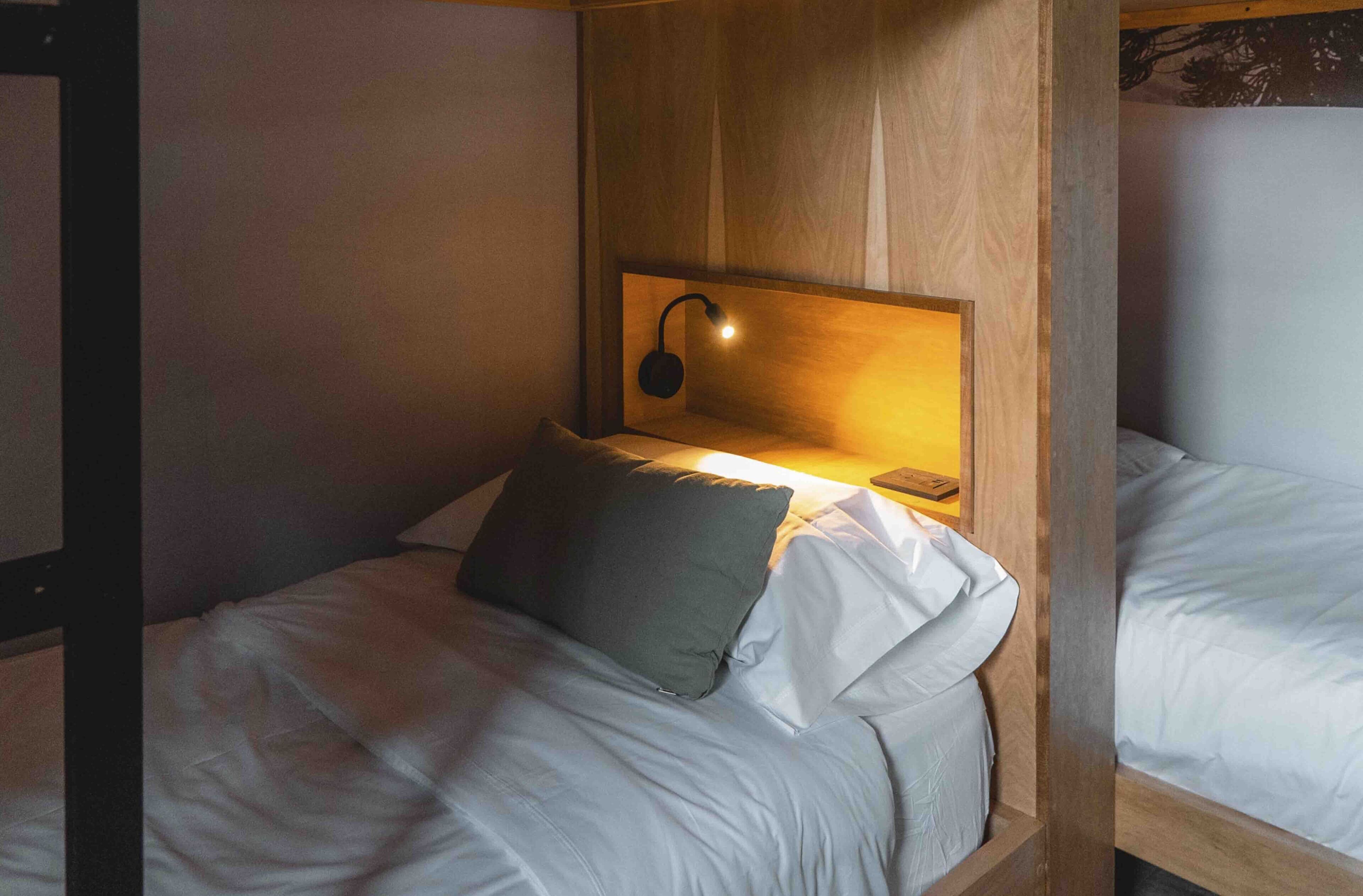 Uma cama beliche com mesa de cabeceira e abajur, roupa de cama branca e cabeceira de madeira.