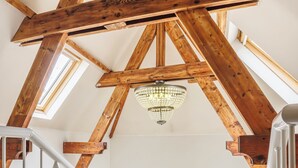 Uma escada de madeira com corrimãos brancos que leva a um teto alto com vigas aparentes e um candelabro.