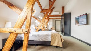 Quarto com teto inclinado, vigas de madeira, cama com roupa de cama branca, mesa de cabeceira, cadeira, escrivaninha, abajur, guarda-roupa e televisão na parede.