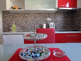 Uma cozinha com um suporte de sobremesa de vidro em dois níveis sobre uma toalha de mesa vermelha, uma cadeira vermelha e um revestimento de azulejos na parede.