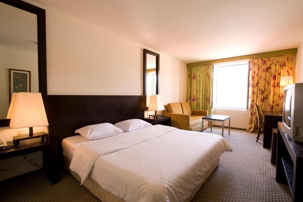 Quarto de hotel com uma cama grande, duas mesas de cabeceira com luminárias, uma cadeira, escrivaninha com televisão e janela com cortinas.