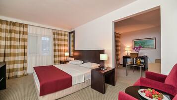 Quarto de hotel com cama, escrivaninha, cadeira, abajur e mesa de jantar com frutas.